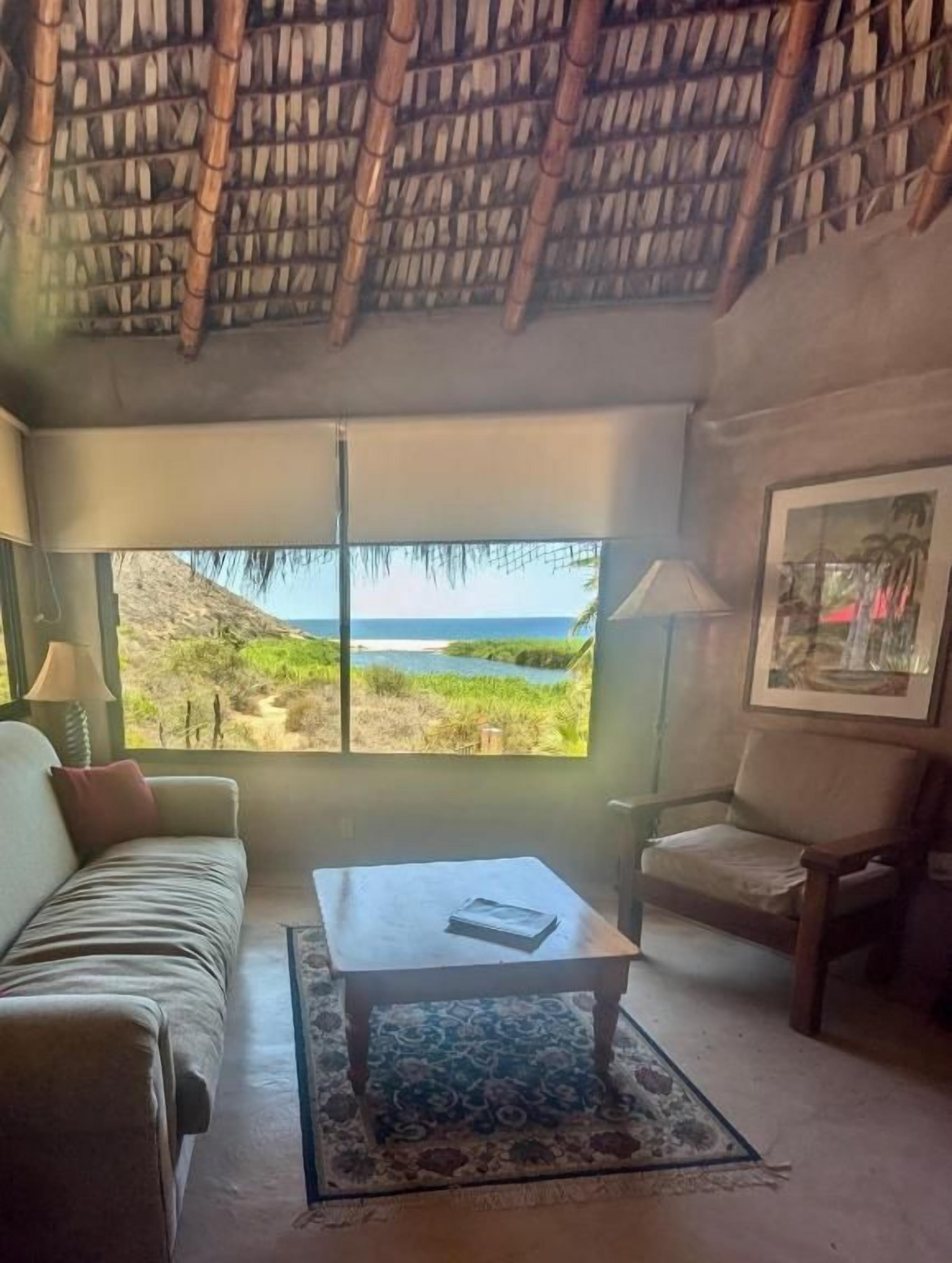 Quarto com vista para a praia e oceano, um sofá, uma cadeira, uma mesa e um abajur.