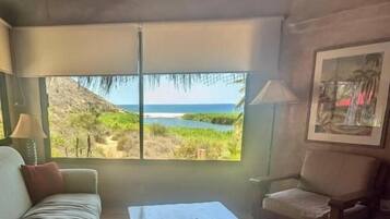 Quarto com vista para a praia e oceano, um sofá, uma cadeira, uma mesa e um abajur.