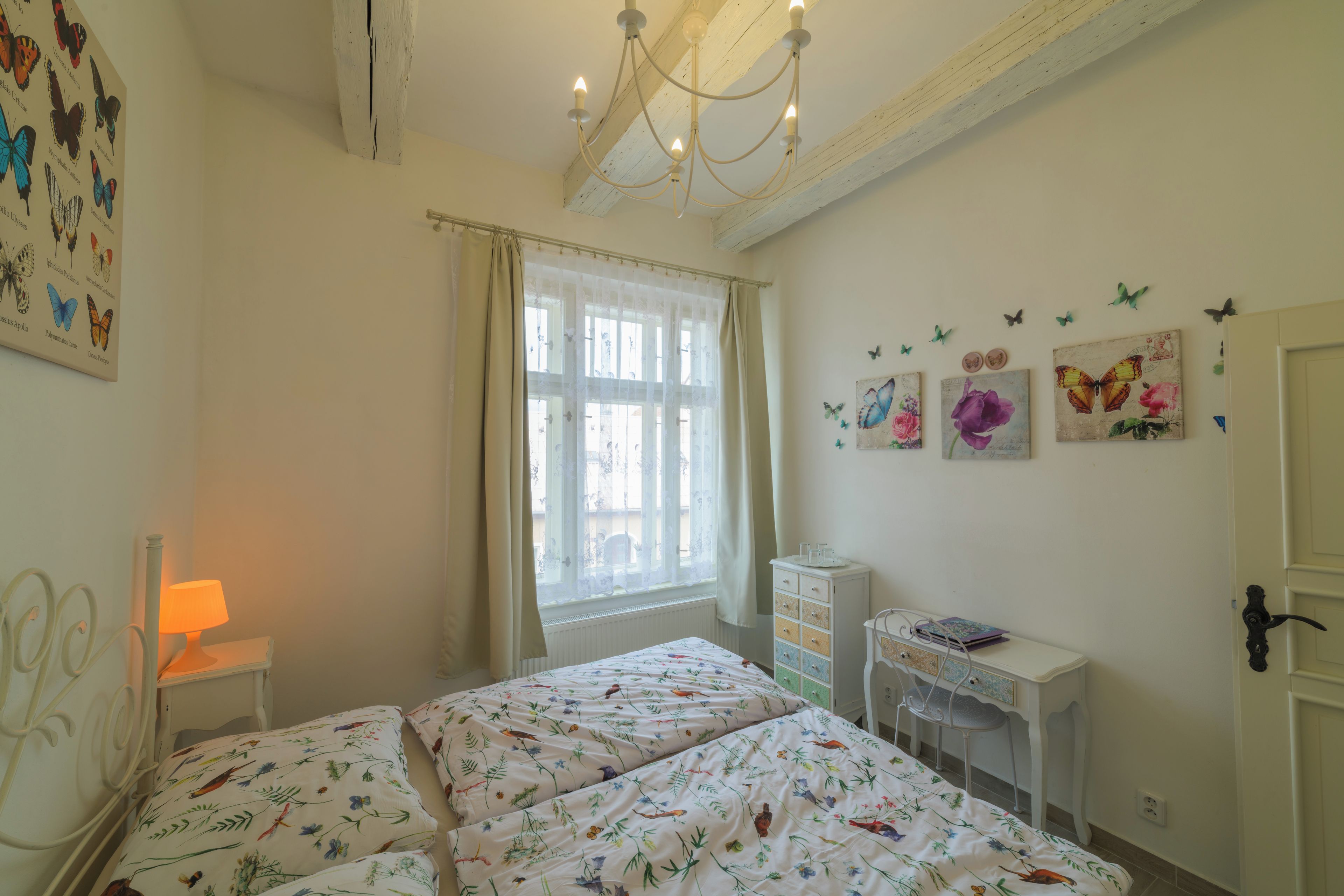 Quarto com cama, mesa de cabeceira, cômoda, janela com cortinas e decorações nas paredes, incluindo borboletas e flores.
