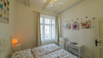 Quarto com cama, mesa de cabeceira, cômoda, janela com cortinas e decorações nas paredes, incluindo borboletas e flores.