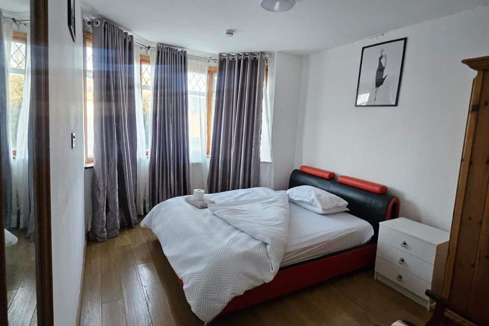 Quarto com cama, mesa de cabeceira, cômoda e janela com cortinas.