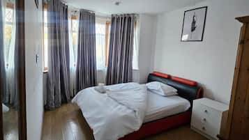 Quarto com cama, mesa de cabeceira, cômoda e janela com cortinas.