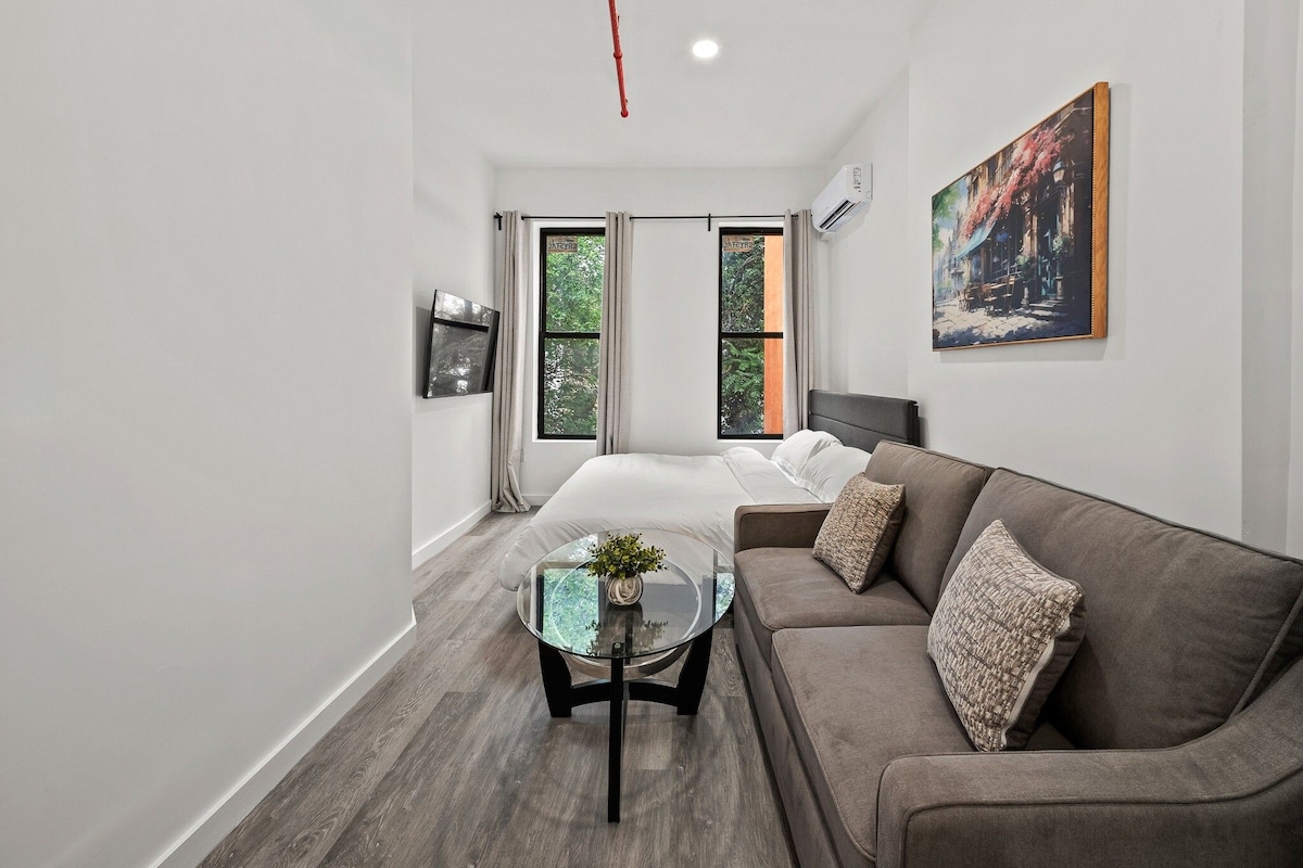 Quarto de hotel moderno com cama, sofá, mesa de centro de vidro e um quadro na parede.