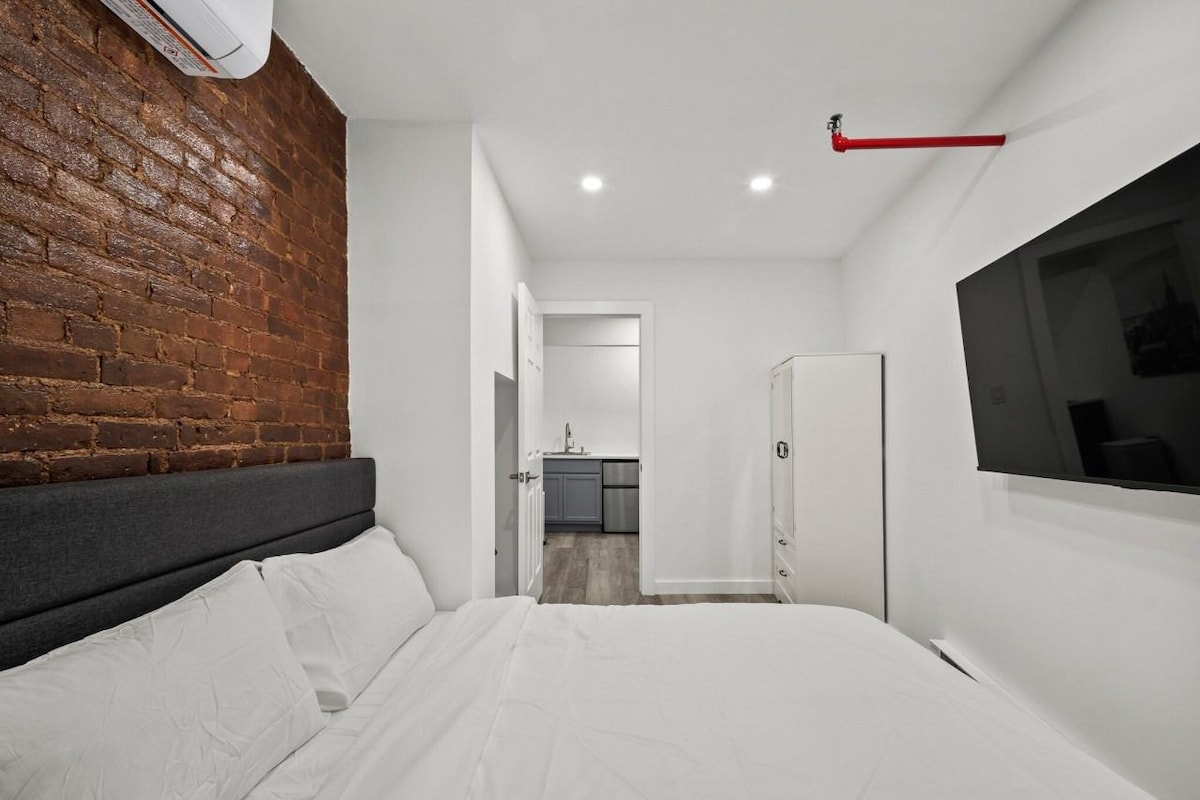 Quarto moderno com parede de tijolos, cama com roupa de cama branca, televisão de tela plana e uma cozinha compacta ao fundo.