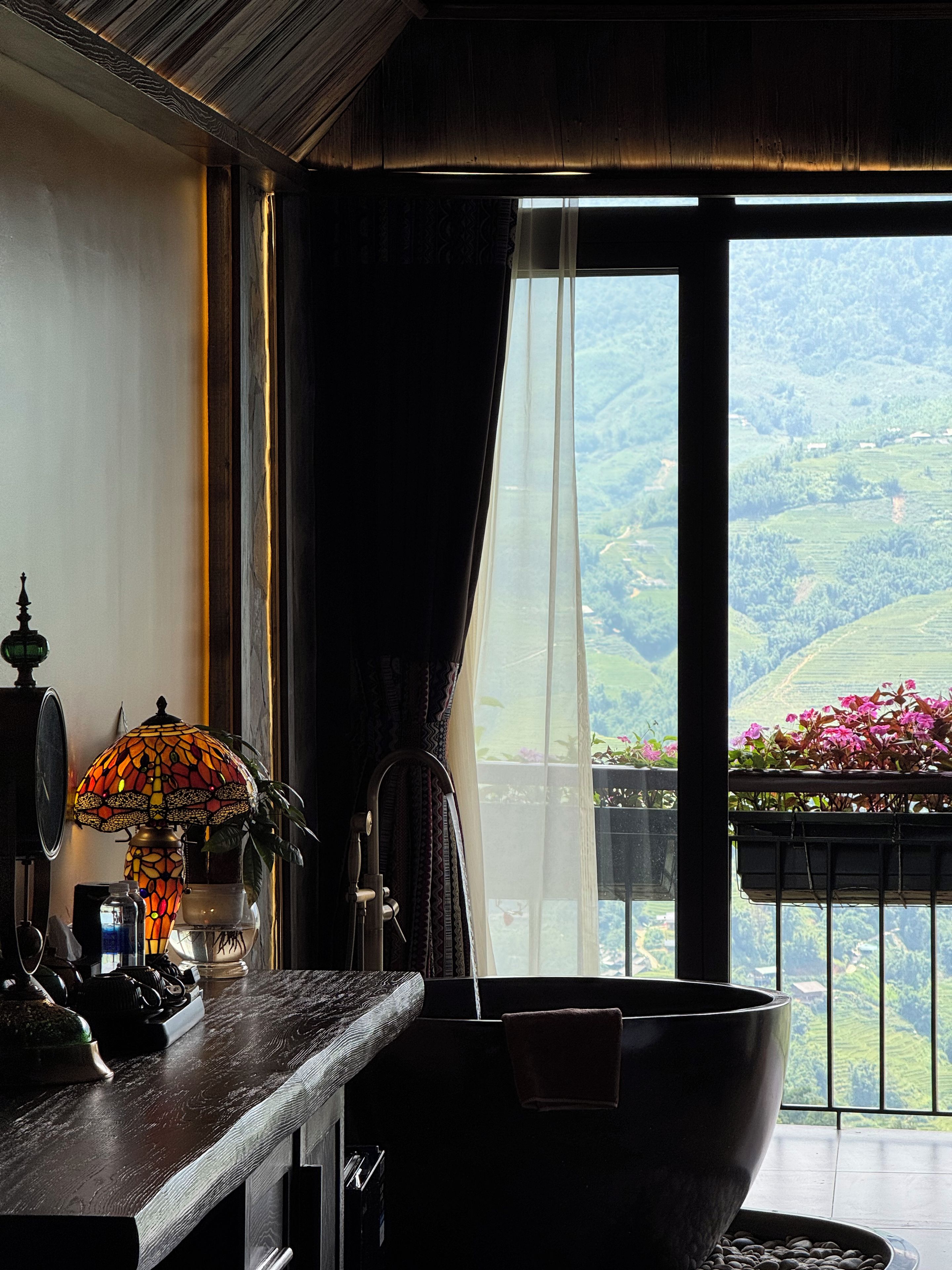 Quarto com uma grande janela, uma escrivaninha de madeira, uma luminária e vista para colinas verdes.