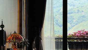 Quarto com uma grande janela, uma escrivaninha de madeira, uma luminária e vista para colinas verdes.