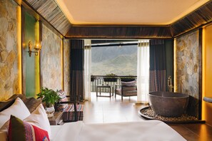 Quarto de hotel com cama grande, banheira, varanda e vista para as montanhas, além de área de jantar.