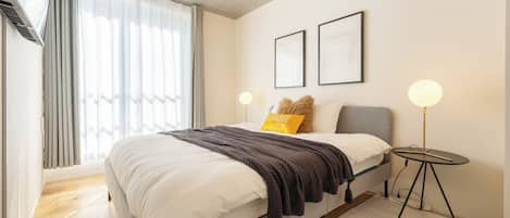 Quarto moderno com cama, mesas de cabeceira, uma janela grande com cortinas e duas imagens emolduradas na parede.