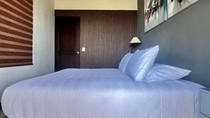 Quarto com cama grande, porta, luminária na parede e janela com persianas.