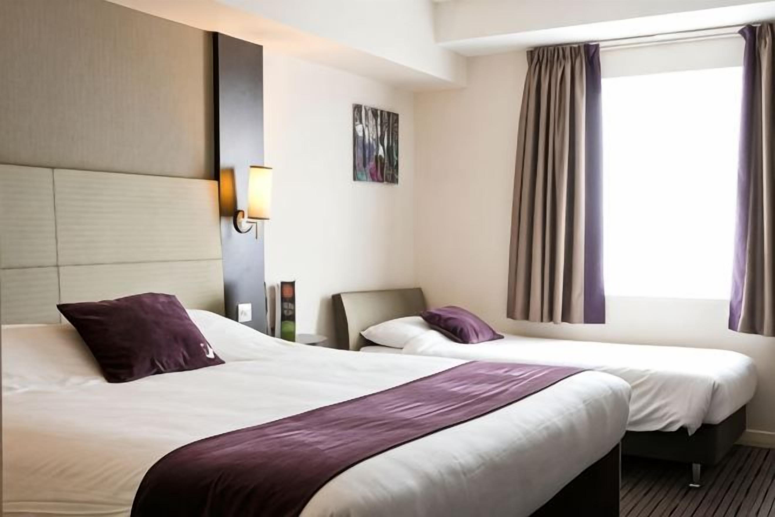 Quarto de hotel com duas camas, cabeceira, mesa de cabeceira, abajur e janela com cortinas.