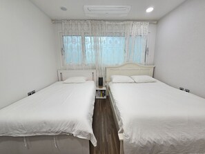 Quarto com duas camas de solteiro, janela com cortinas finas, mesa de cabeceira com livros e ar condicionado no teto.