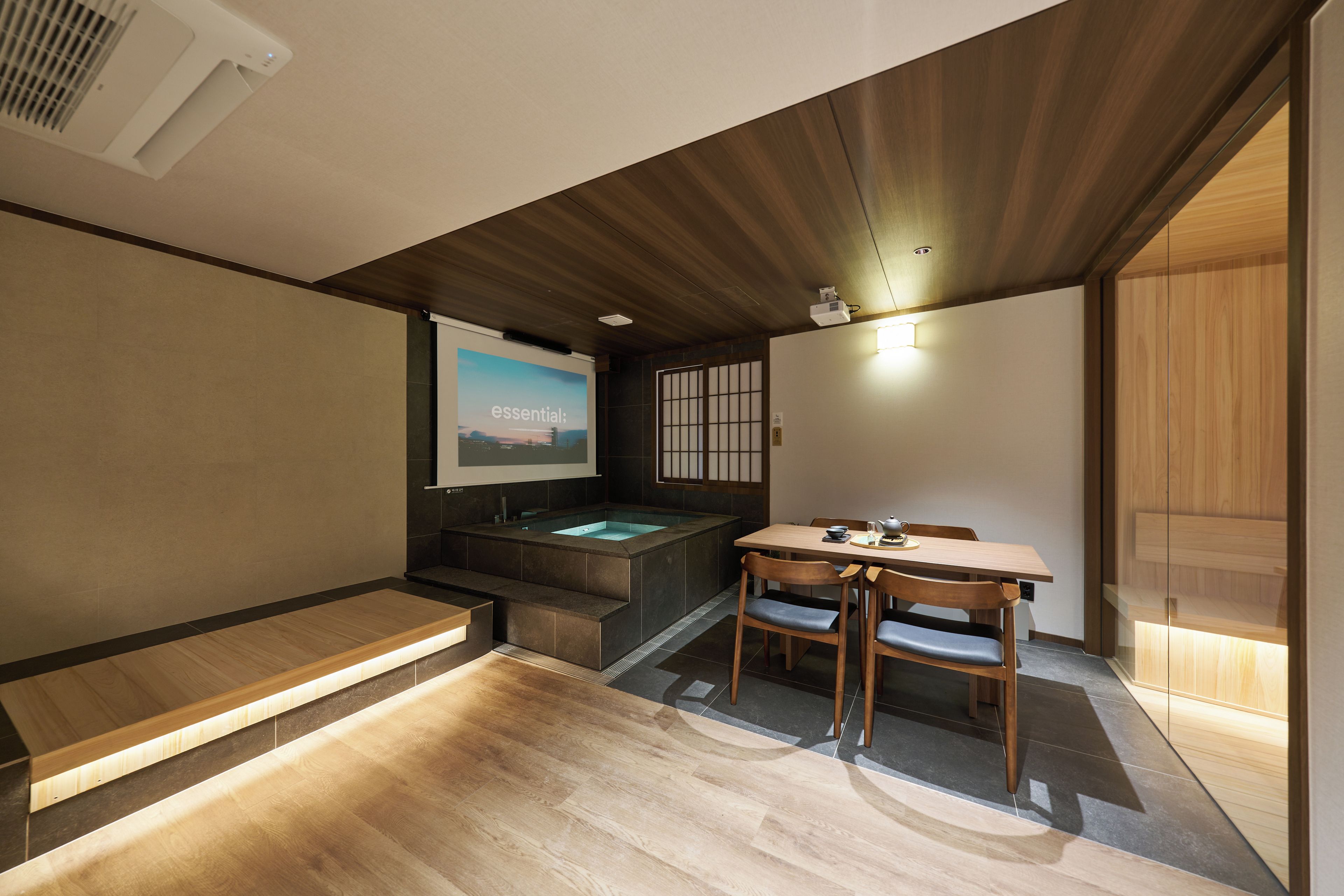 Um interior moderno com piso de madeira, área de estar com mesa e cadeiras, uma pequena banheira de hidromassagem e uma tela de projeção exibindo a palavra 'essential'.