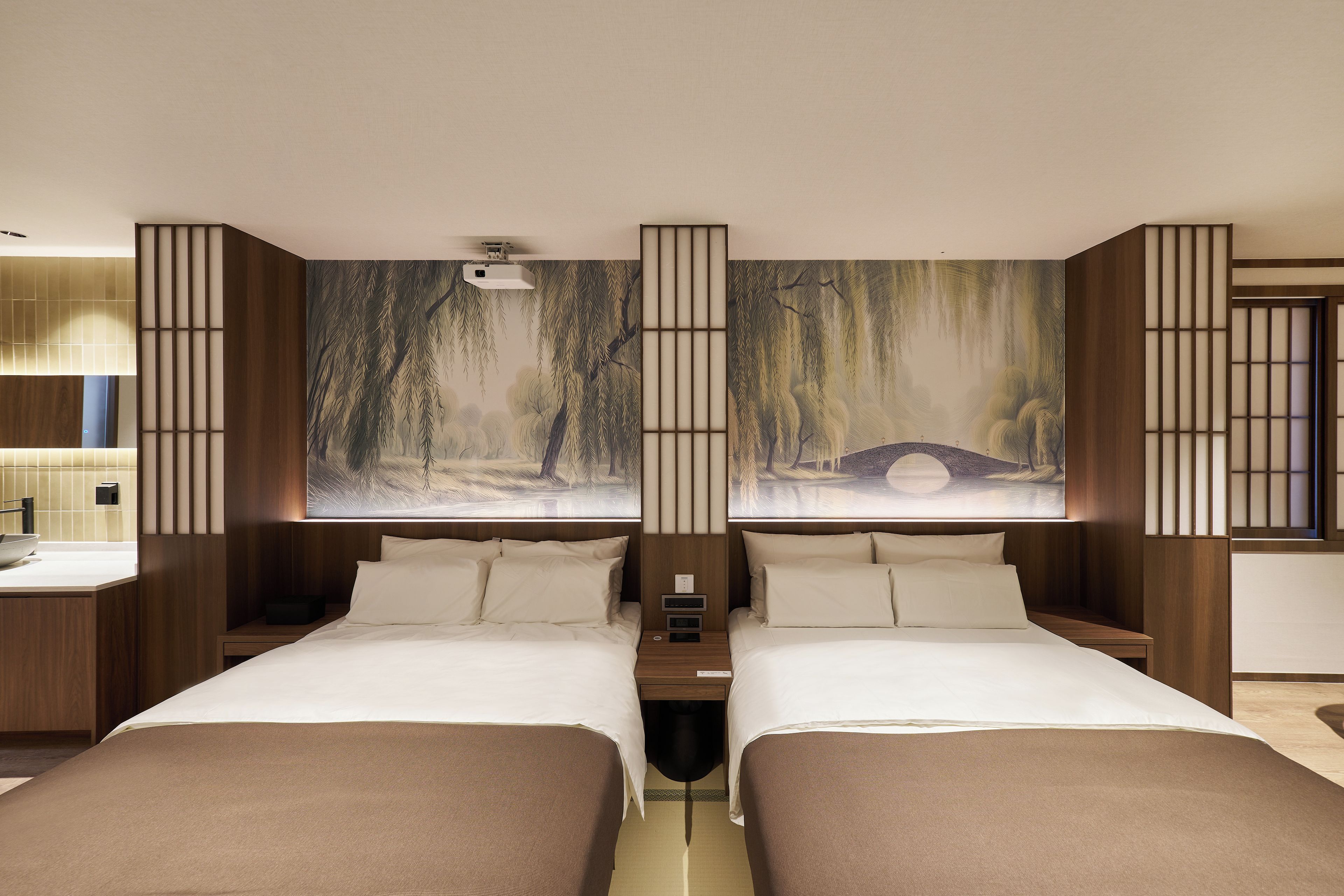 Quarto de hotel com duas camas, um grande mural na parede, projetor no teto e banheiro moderno.