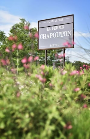Placa indicando 'Hostellerie La Ferme Chapouton Bistro', com vista nítida da vegetação ao redor e uma estrutura de estufa ao fundo.