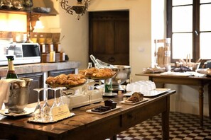 Mesa de buffet com diversos alimentos, uma garrafa de champanhe e uma máquina de café ao fundo.