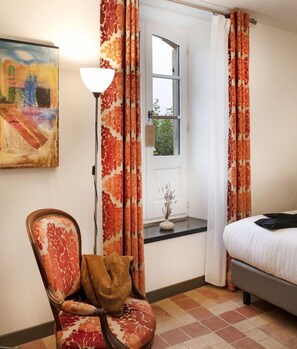 Quarto de hotel com cama, cadeira com almofada, luminária e janela com cortinas.