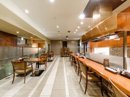 Uma sala de jantar de hotel com mesas e cadeiras de madeira, uma área de cozinha com eletrodomésticos de aço inoxidável e piso ladrilhado.