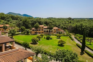 Um edifício grande, com vários andares e um pátio central, rodeado por vegetação exuberante e colinas ao fundo.
