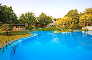 Piscina com superfície de água azul cristalina, rodeada por árvores e um edifício ao fundo.