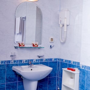 Casa de banho com uma pia branca, espelho, suporte de toalhas, prateleira de vidro, porta-papel higiénico e telefone fixado na parede.