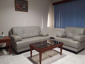 Uma sala de estar com dois sofás, uma mesa de centro, um quadro na parede e uma janela com persianas.