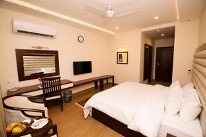 Quarto de hotel com cama, escrivaninha com cadeira, televisão e relógio na parede.