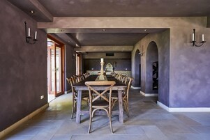 Uma sala de jantar com uma mesa de madeira comprida e cadeiras, um candelabro e vista para a cozinha.