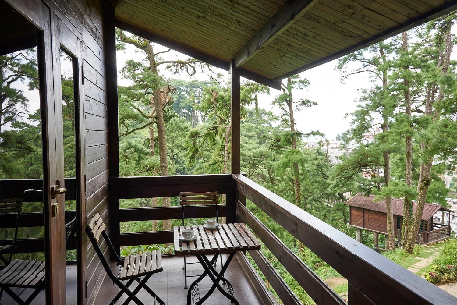 Um deck de madeira com um banco e uma mesa, com vista para uma área arborizada com árvores e um pequeno edifício ao fundo.