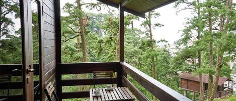 Um deck de madeira com um banco e uma mesa, com vista para uma área arborizada com árvores e um pequeno edifício ao fundo.