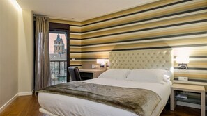 Quarto de hotel com uma cama grande, uma mesa de cabeceira, abajur, uma cadeira e vista para um prédio pela janela.