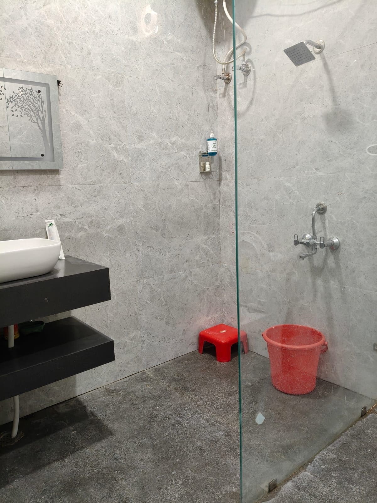 Casa de banho com lavatório branco, cabine de duche em vidro, um balde vermelho e um taburete pequeno.