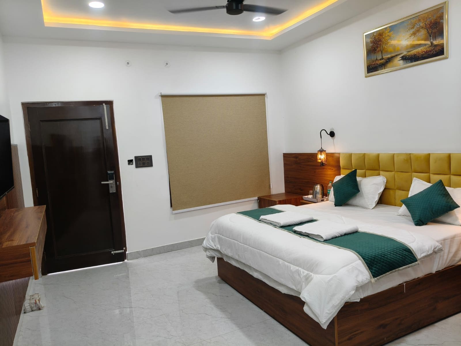 Quarto de hotel com cama, mesa de cabeceira, ventoinha no teto, um quadro na parede e uma porta.