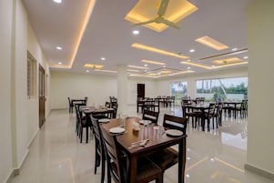 Uma sala de jantar ampla com várias mesas e cadeiras, ventiladores de teto, iluminação embutida e grandes janelas.
