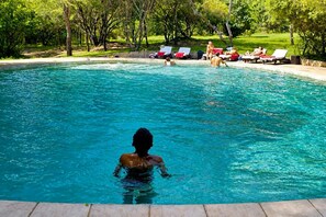 Piscina com água azul cristalina, rodeada por espreguiçadeiras e um gramado.