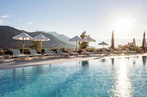 Área de piscina com espreguiçadeiras e guarda-sóis, céu claro e montanhas ao fundo.