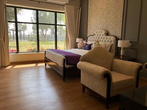 Quarto com cama, cadeira, janela com cortinas e vista para um jardim.