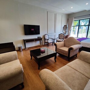 Quarto de hotel com TV de tela plana, sofá, mesa de centro e janela com vista para áreas verdes.