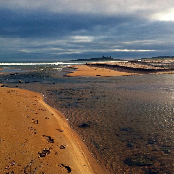 Uma praia com pegadas na areia, uma construção distante e ondas se aproximando da costa.