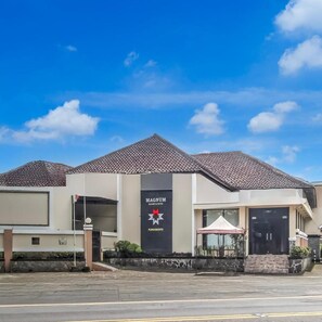 Um edifício com telhado em cerâmica, um grande letreiro com o nome 'MAGNUM RESORT & HOTEL' e um céu azul claro ao fundo.