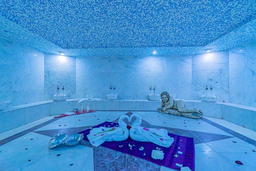 Um espaço de spa com piso de mármore, área central com toalhas dispostas em formato de coração e uma estátua de uma divindade.
