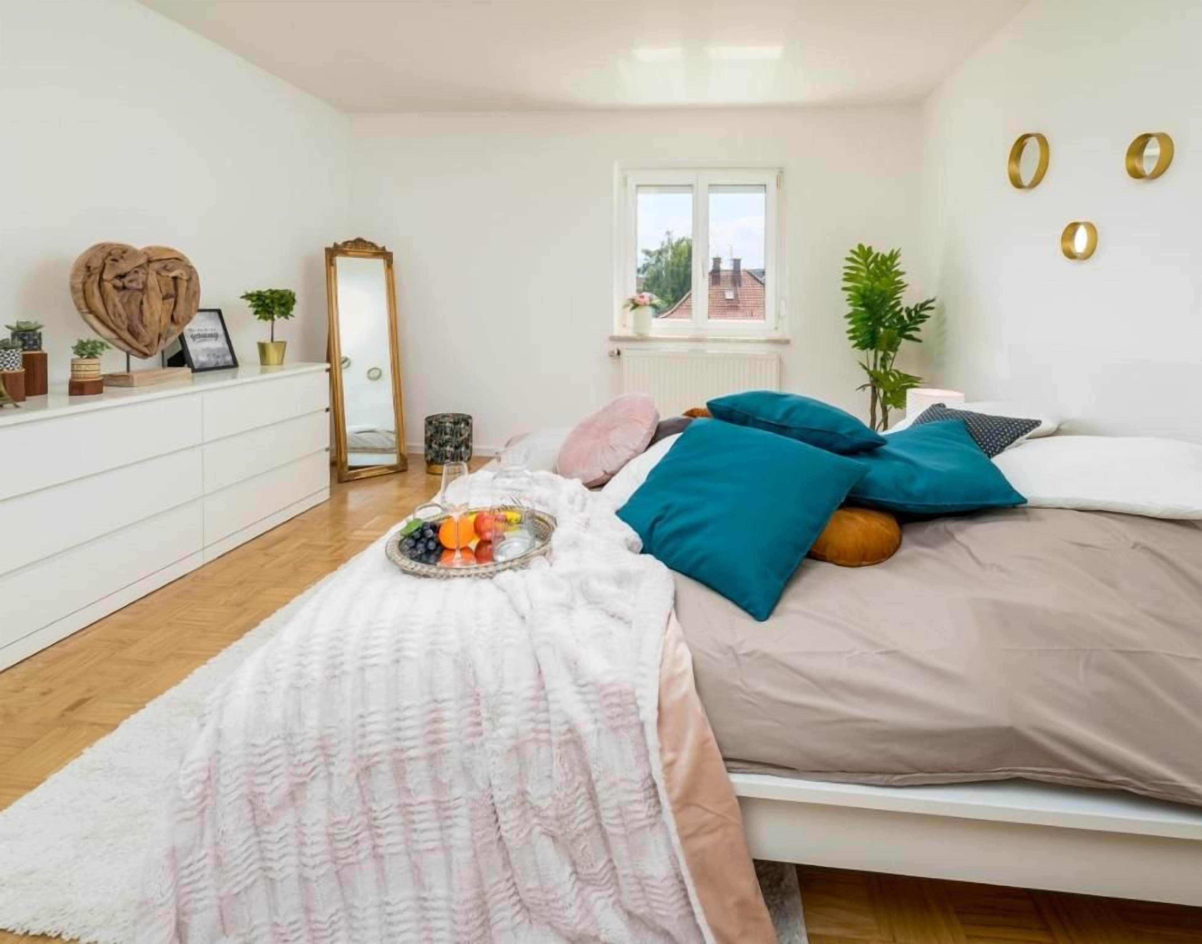 Quarto com cama, mesa de cabeceira, cômoda, espelho, janela e uma planta.