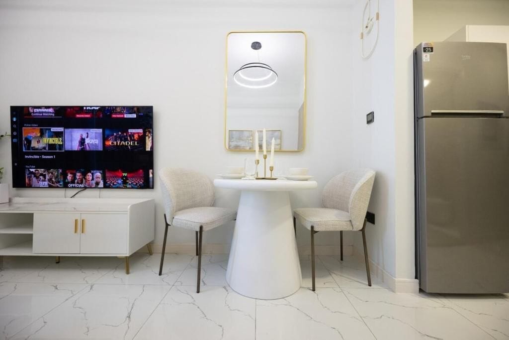 Uma sala de estar moderna com televisão de tela plana, uma mesa de jantar branca com duas cadeiras, uma geladeira e um luminário fixado na parede.