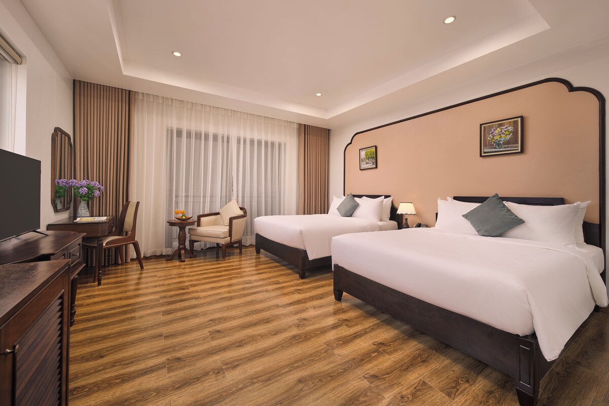 Quarto de hotel com duas camas, uma escrivaninha com cadeira, televisão e janela com cortinas.