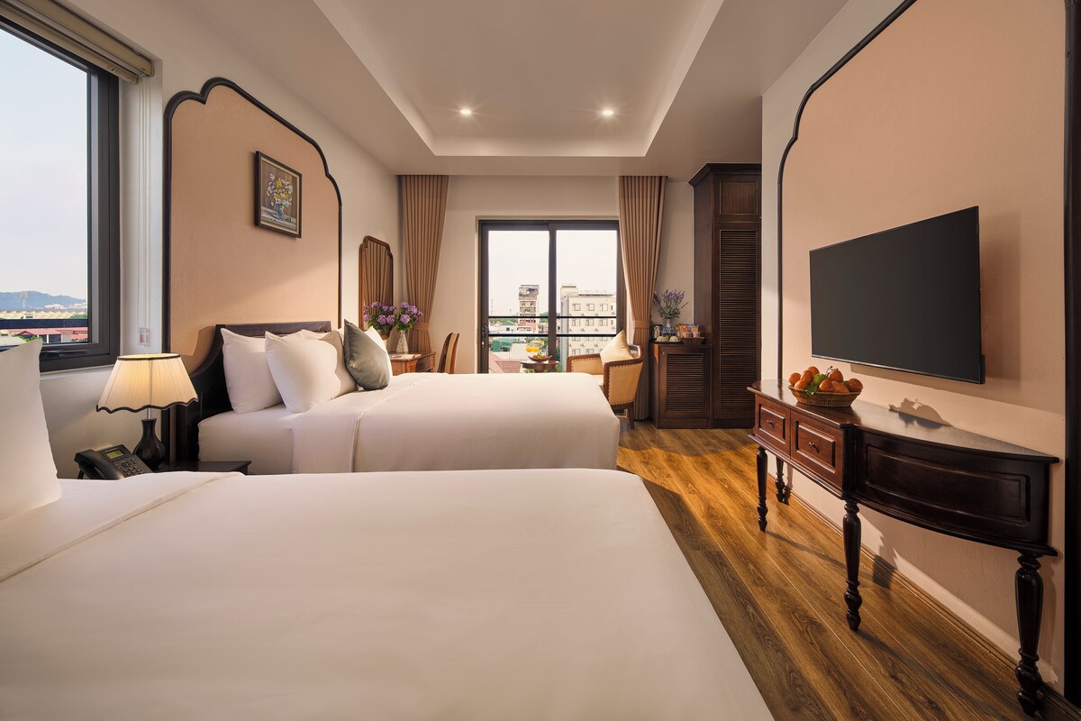 Quarto de hotel com duas camas, uma escrivaninha, televisão e varanda com vista.