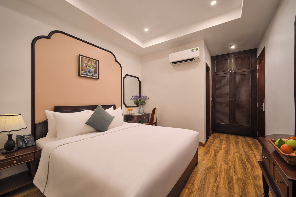 Quarto de hotel com uma cama grande, mesa de cabeceira com luminária, telefone, espelho, ar condicionado, guarda-roupa de madeira e cesta de frutas.