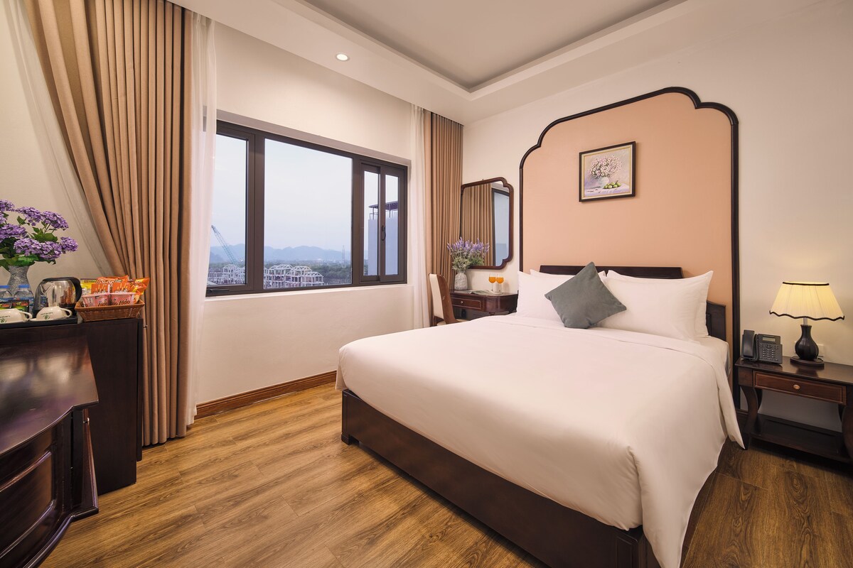 Quarto de hotel com uma cama grande, uma escrivaninha com abajur, telefone e janela com cortinas.