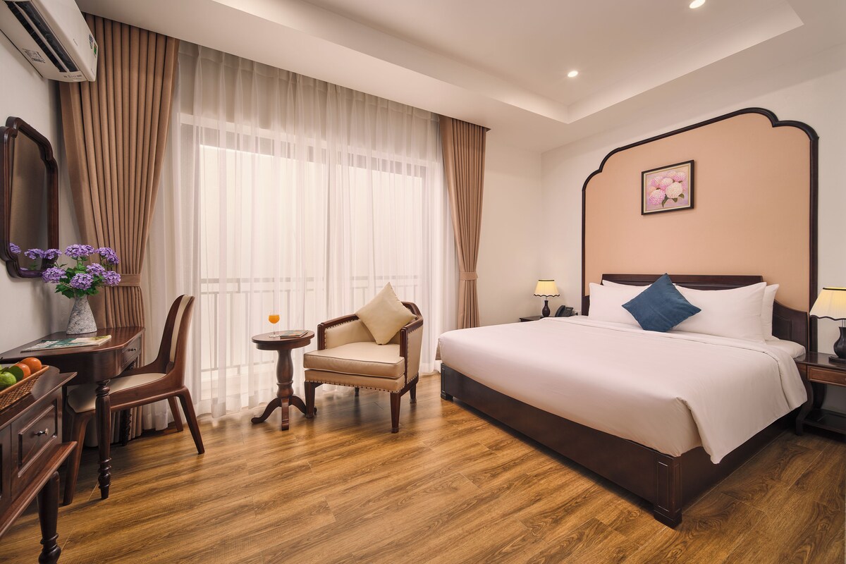 Quarto de hotel com uma cama grande, uma escrivaninha com cadeira, uma mesinha de cabeceira com abajur e uma janela com cortinas.