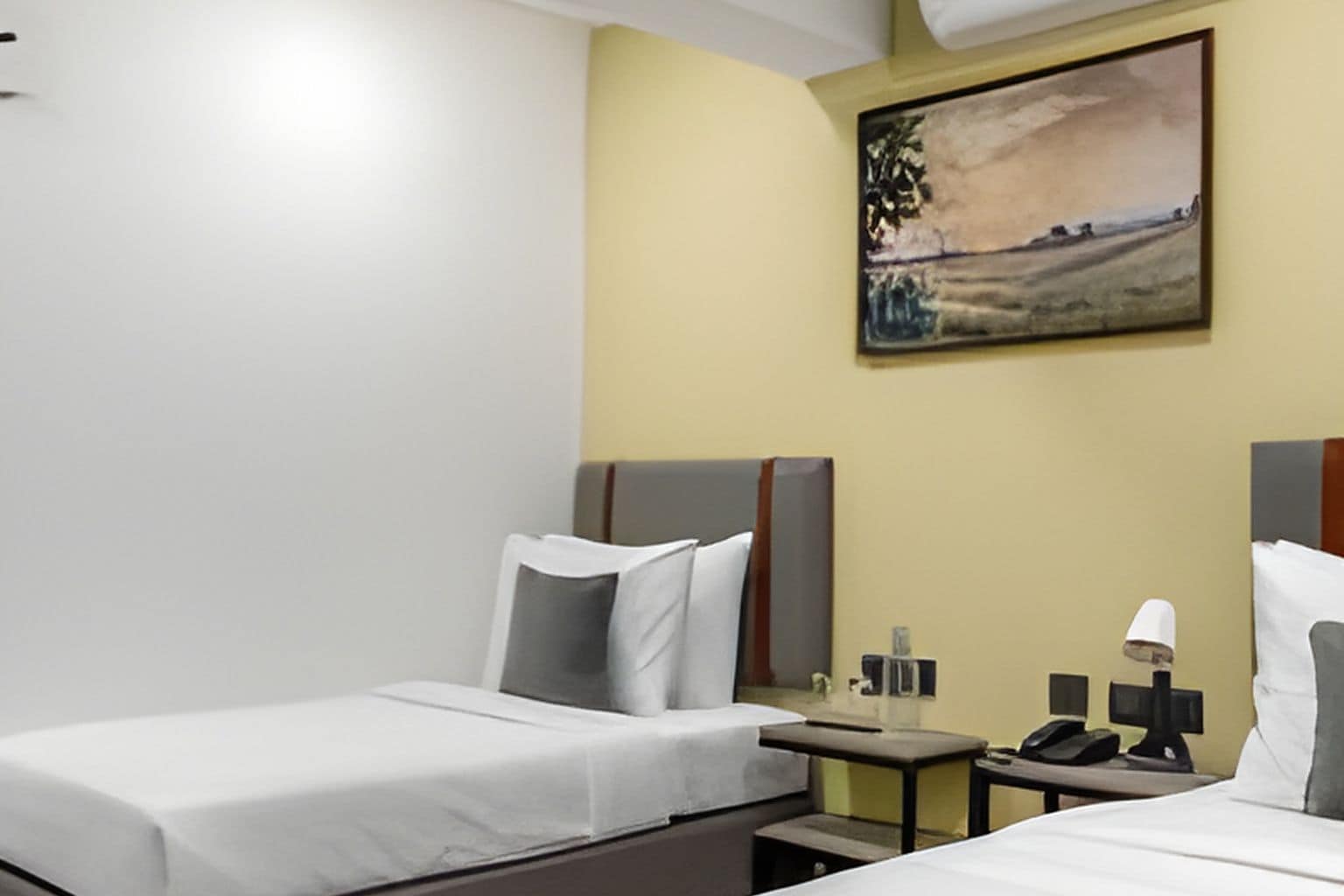Quarto de hotel com duas camas, uma mesa de cabeceira com abajur, um quadro na parede e uma pequena mesa com telefone.