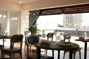 Uma área de refeições no terraço com mesas e cadeiras, vista para a paisagem urbana e uma parede de vidro.