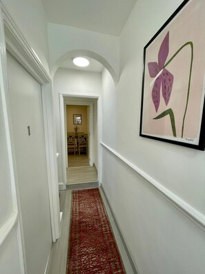 Um corredor com carpete vermelho estampado, paredes brancas e uma pintura emoldurada de uma flor roxa.